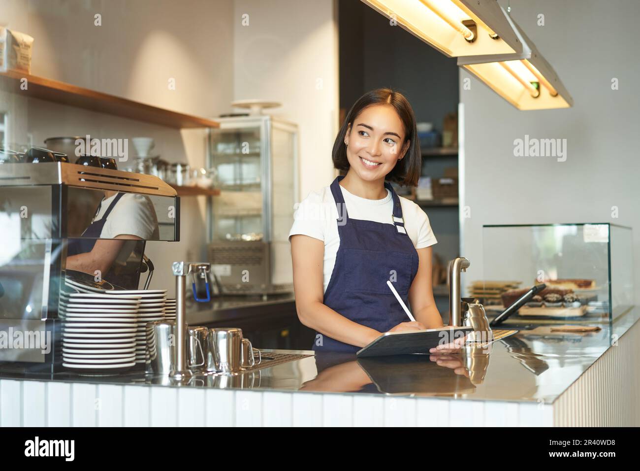 Portrait of beautiful asian girl smiling, barista in cafe working behind counter, using tablet ...