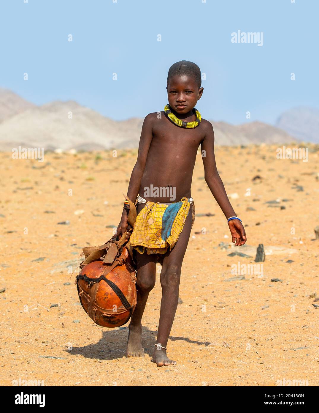 Child of the Himba tribe walks through the desert carrying water in a ...