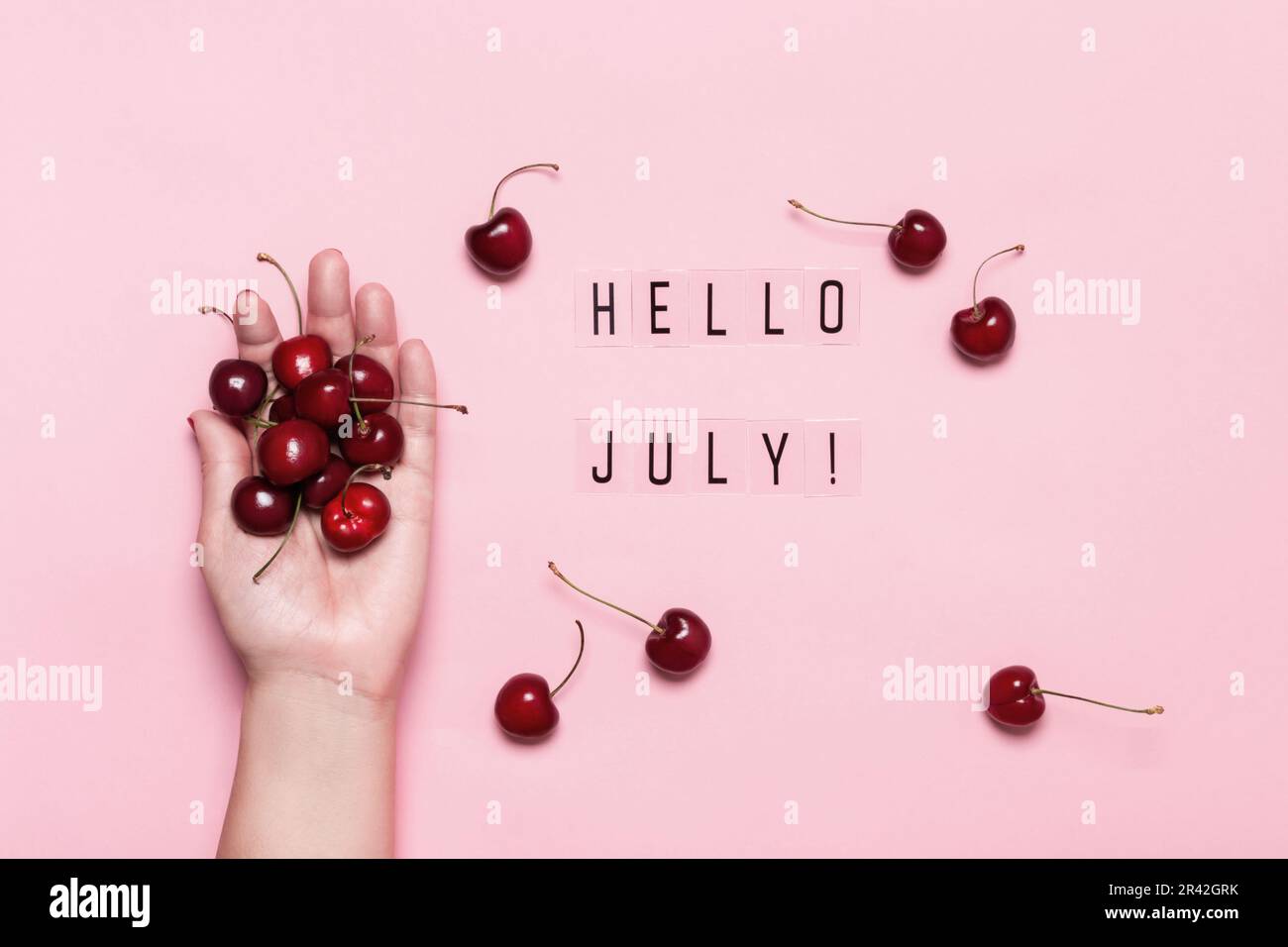 Hello July text and female hand holds ripe cherries on pink background ...