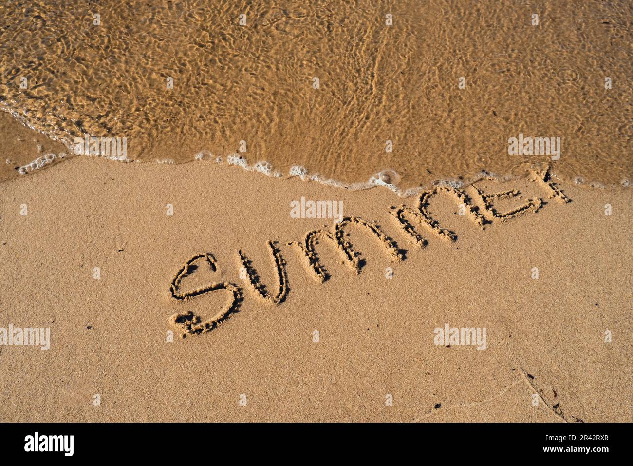 Text SUMMER handwritten in sand surface. Blue ocean wave washing away ...