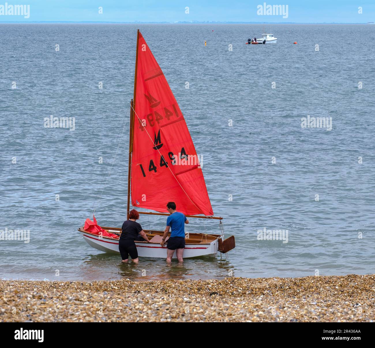 Man & woman climb into a sailing boat with orange sail near the shore ...