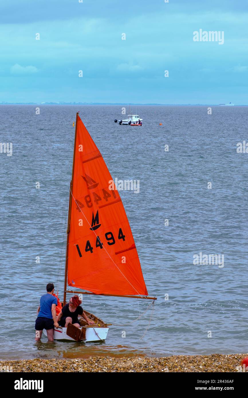 Man & woman climb into a sailing boat with orange sail near the shore ...