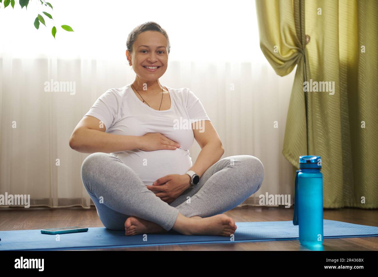 Happy adult pregnant woman gently stroking her belly, sitting in lotus pose on a mat during ...