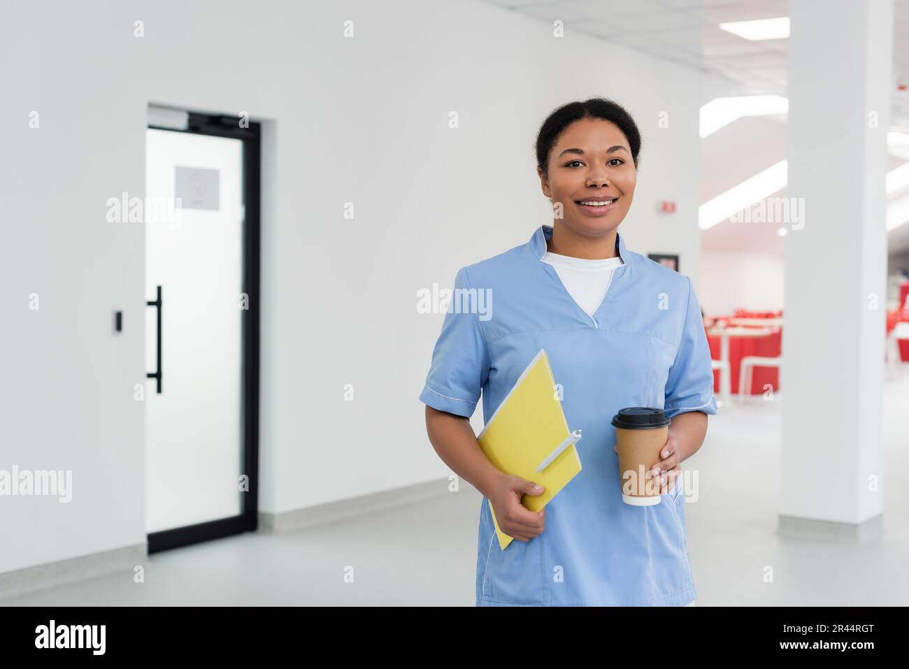 happy multiracial nurse in blue uniform holding folder with pen ...