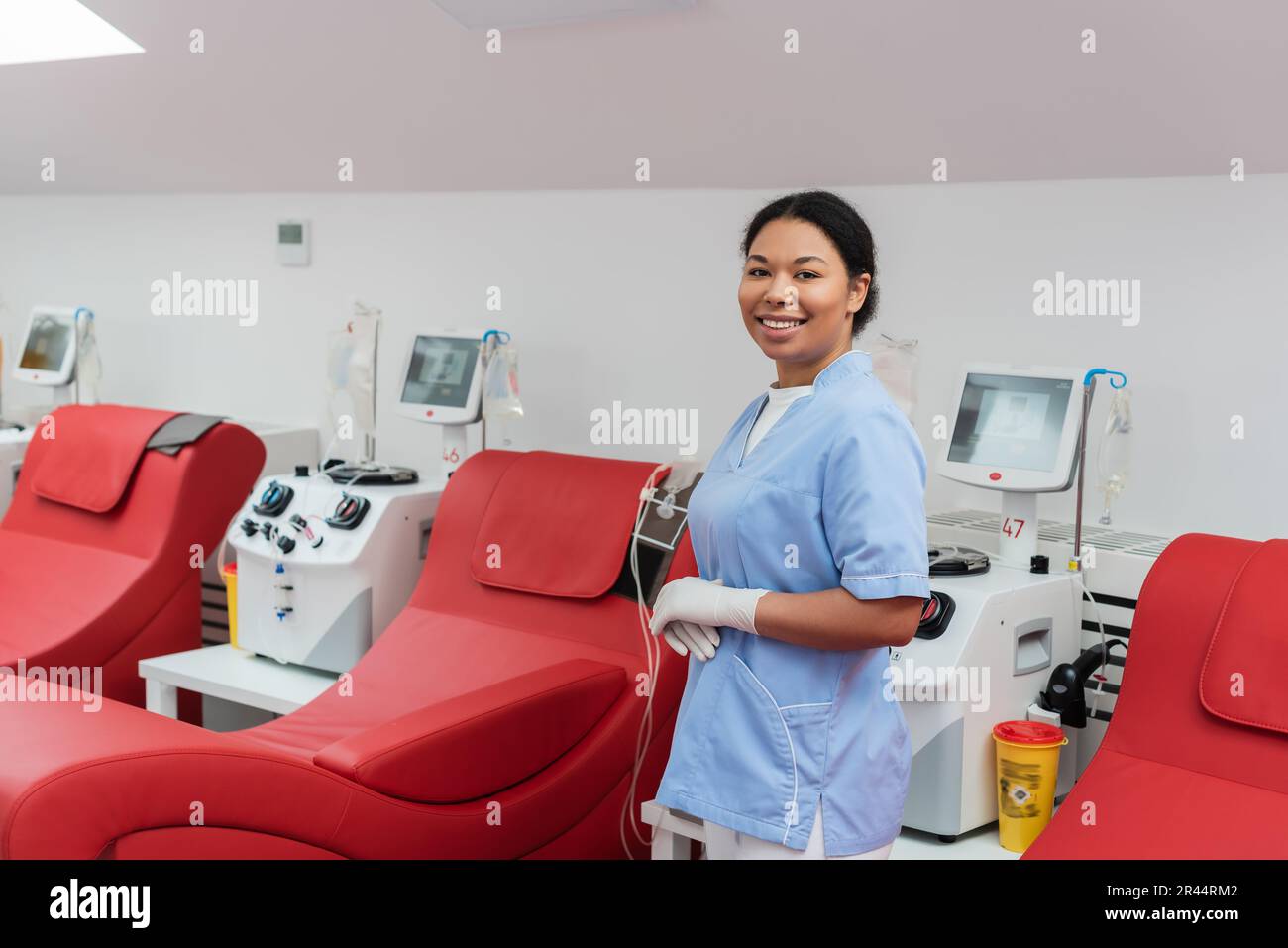 joyful multiracial nurse in blue uniform and latex gloves looking at ...