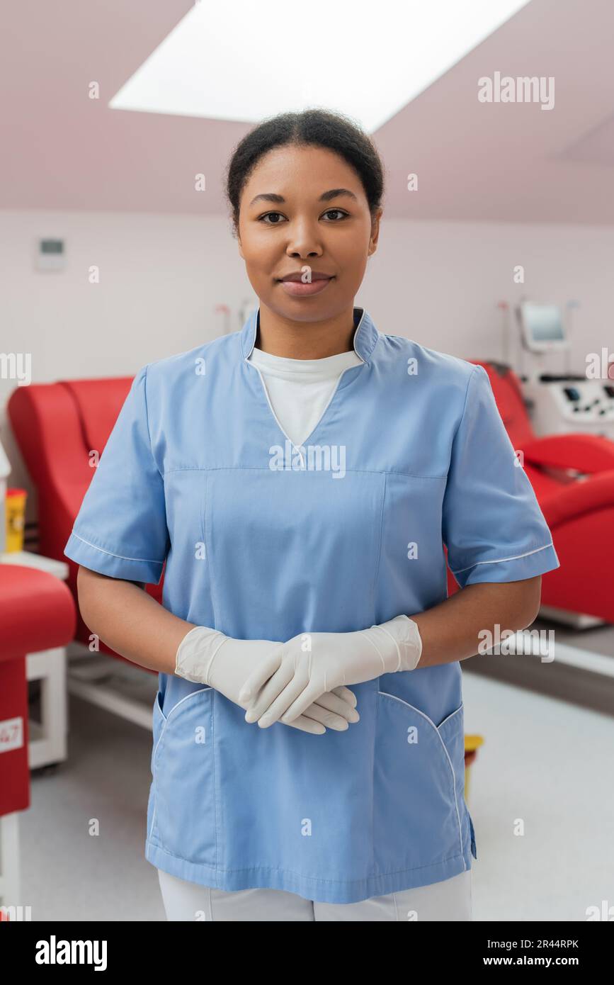 positive multiracial nurse in blue uniform and latex gloves looking at ...