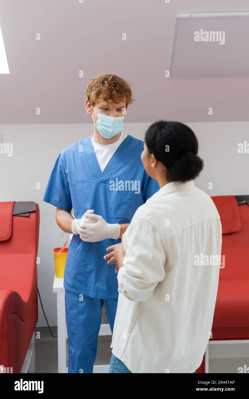 young and redhead doctor in medical mask, latex gloves and blue uniform ...