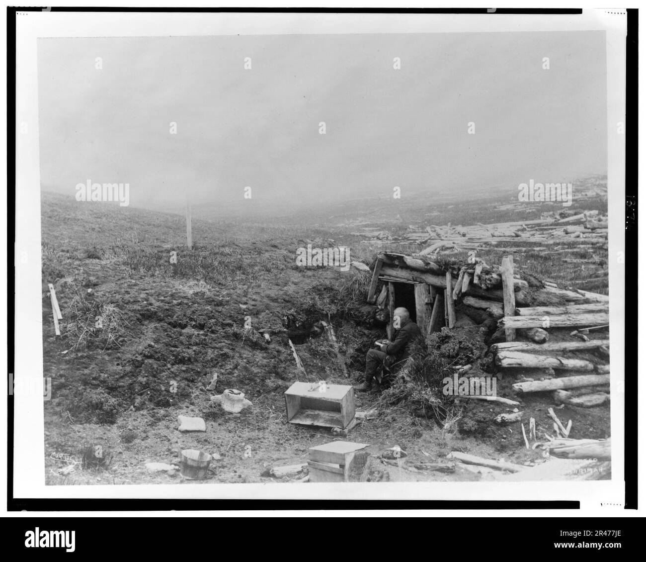 Unidentified old man, seated, writing, at entrance to a log and earth ...