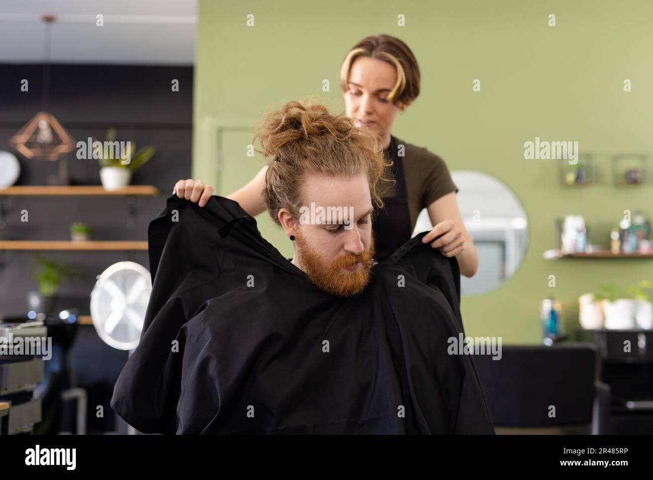 Caucasian female barber putting cape on male customer at barbershop ...