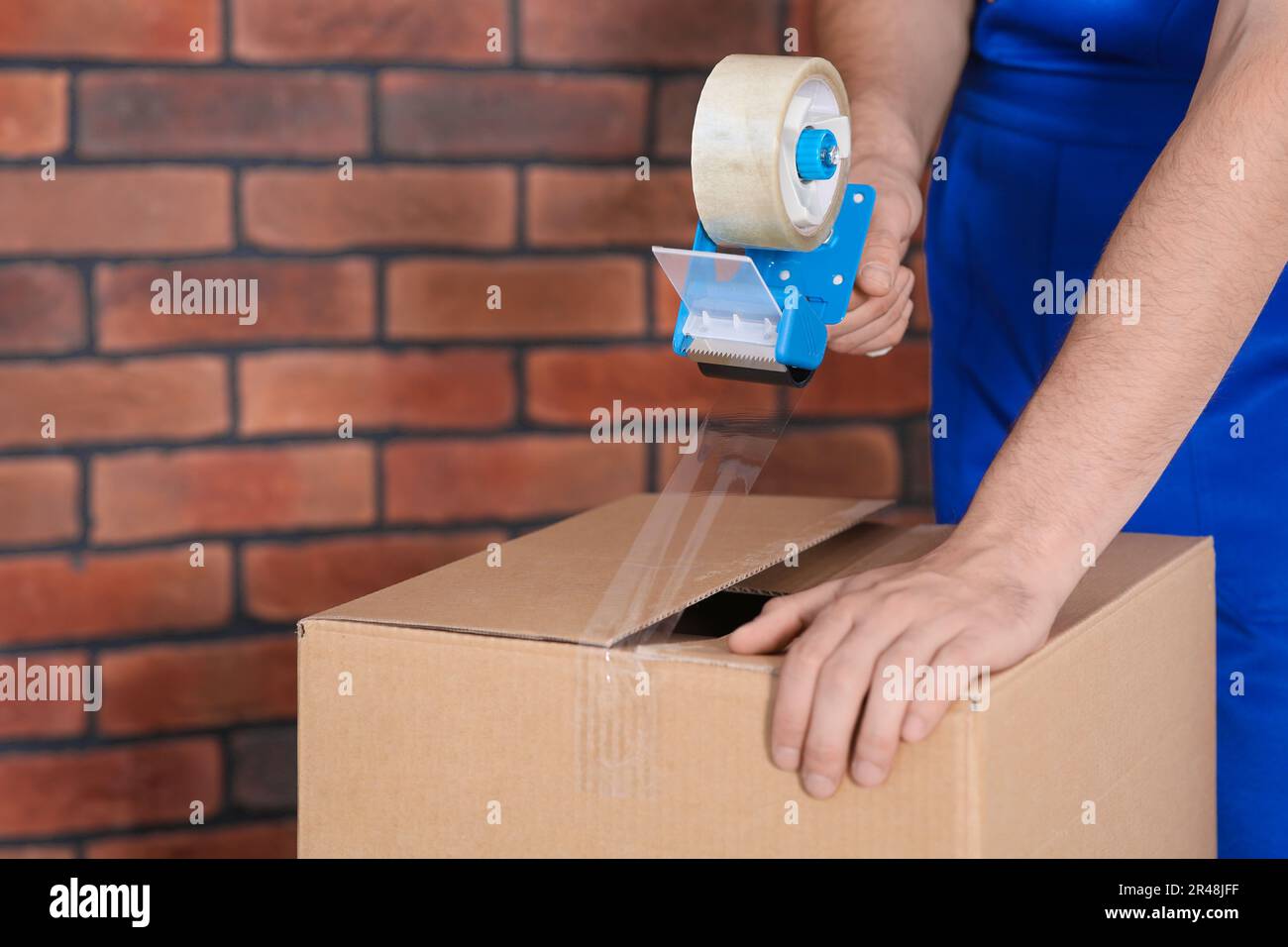 Worker taping box with adhesive tape dispenser near brick wall, closeup ...
