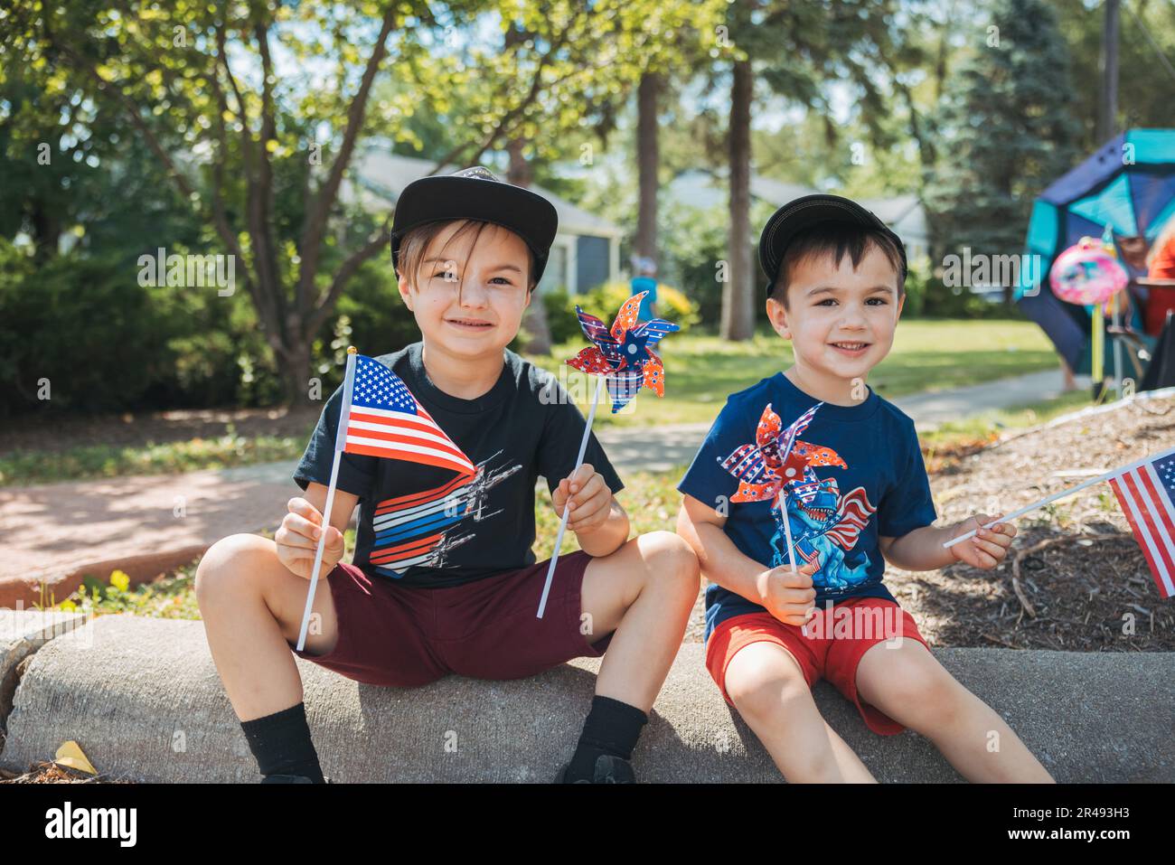Two young boys holding US flags sitting on a park bench, smiling and ...