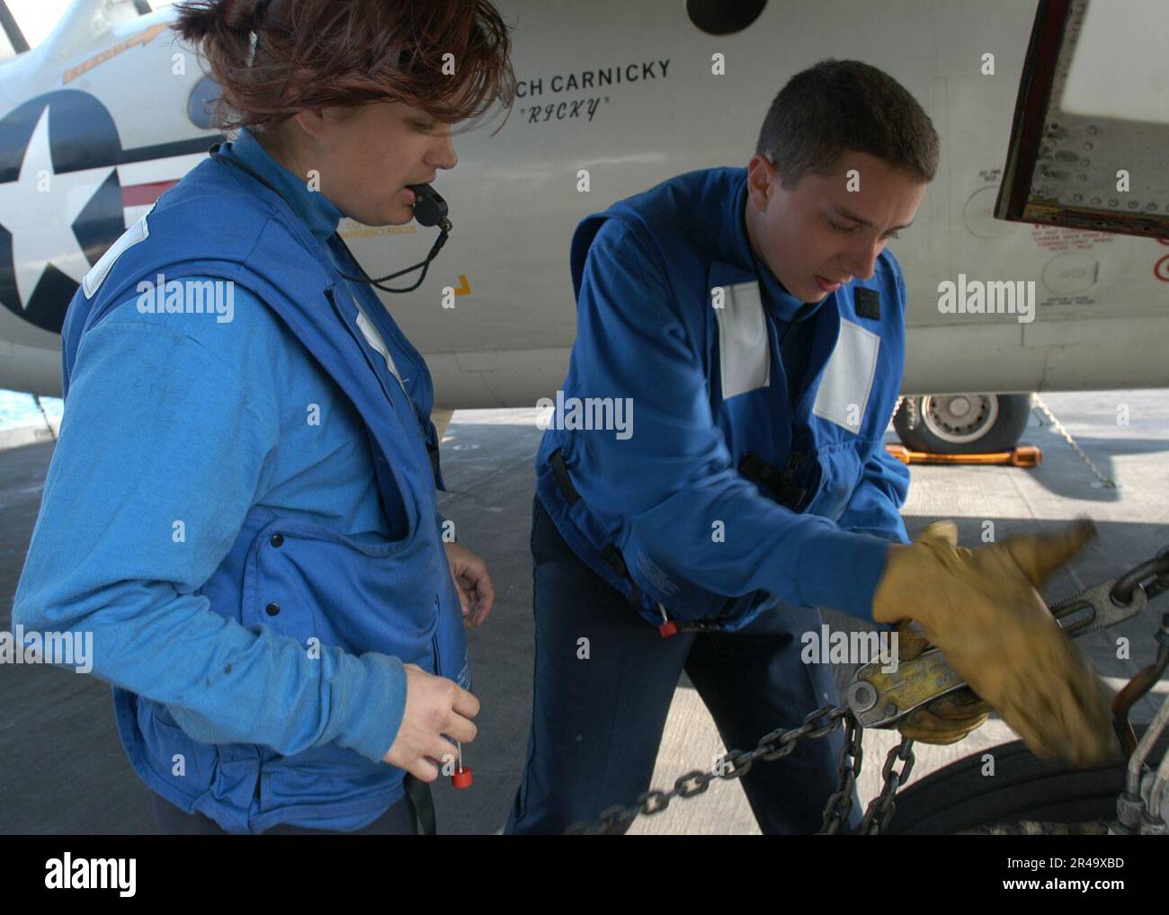 US Navy Airman supervises Airman as he uses a tie-down chain to secure ...