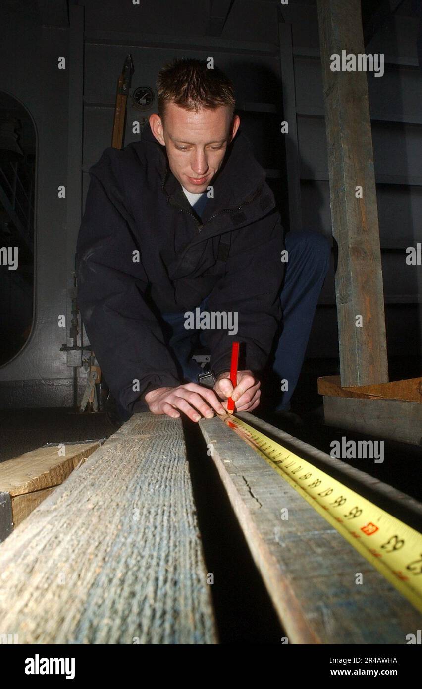 US Navy Damage Controlman 3rd Class measures wood before cutting it ...