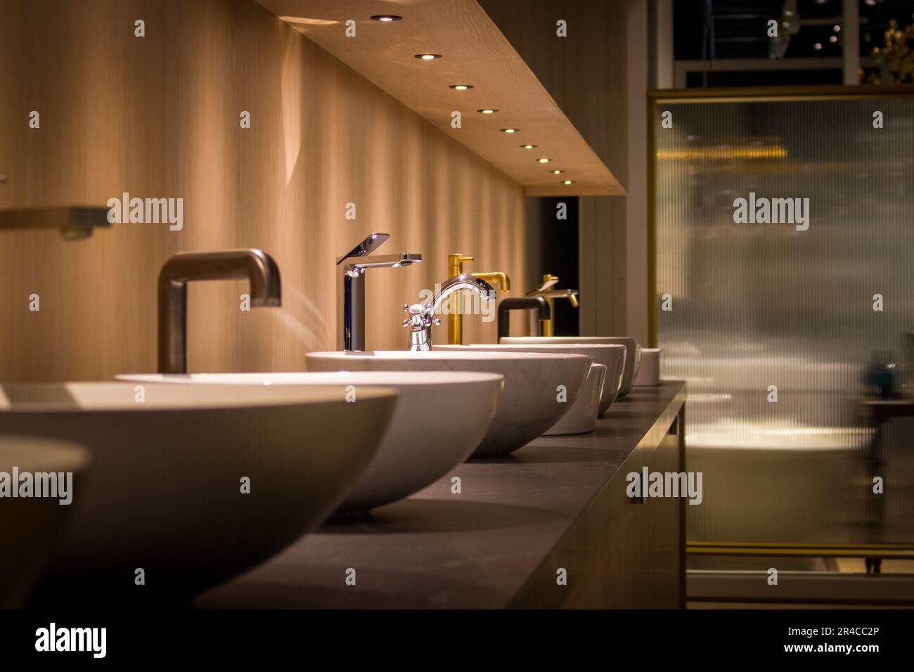This image depicts a modern restroom with six white ceramic basins, one ...