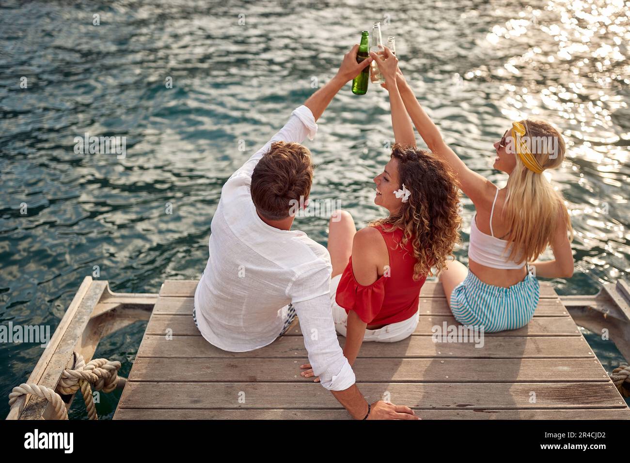 Group of friends toasting with beer chilling on wooden jetty by water ...