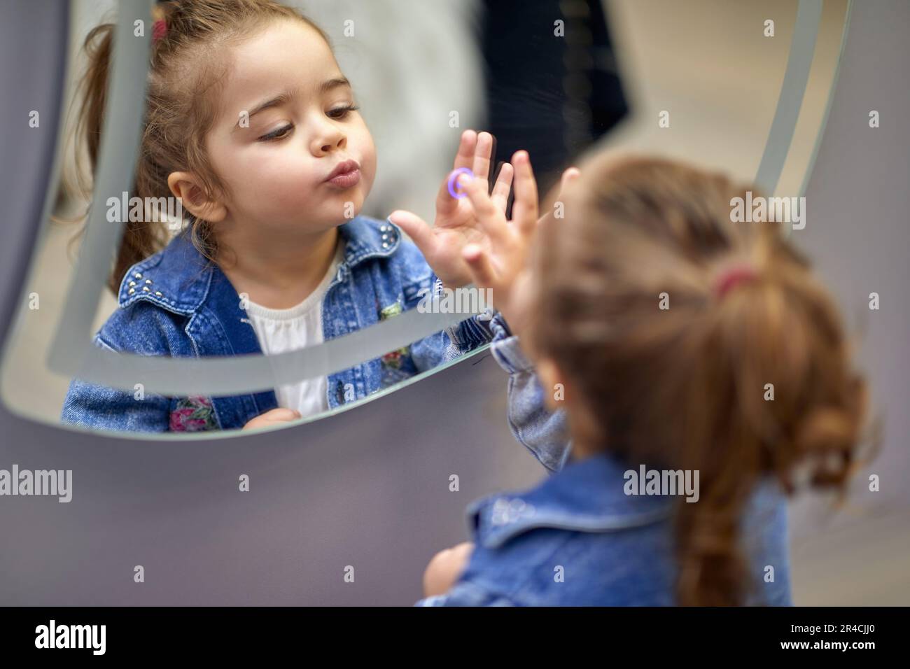 Adorable little girl making faces and looking at herself in the mirror ...