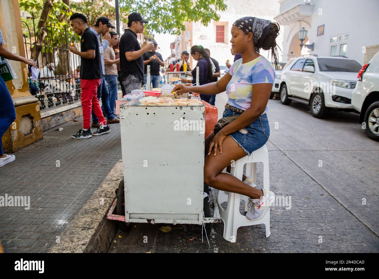 A young woman sits at an arepas street food stand while people stand ...