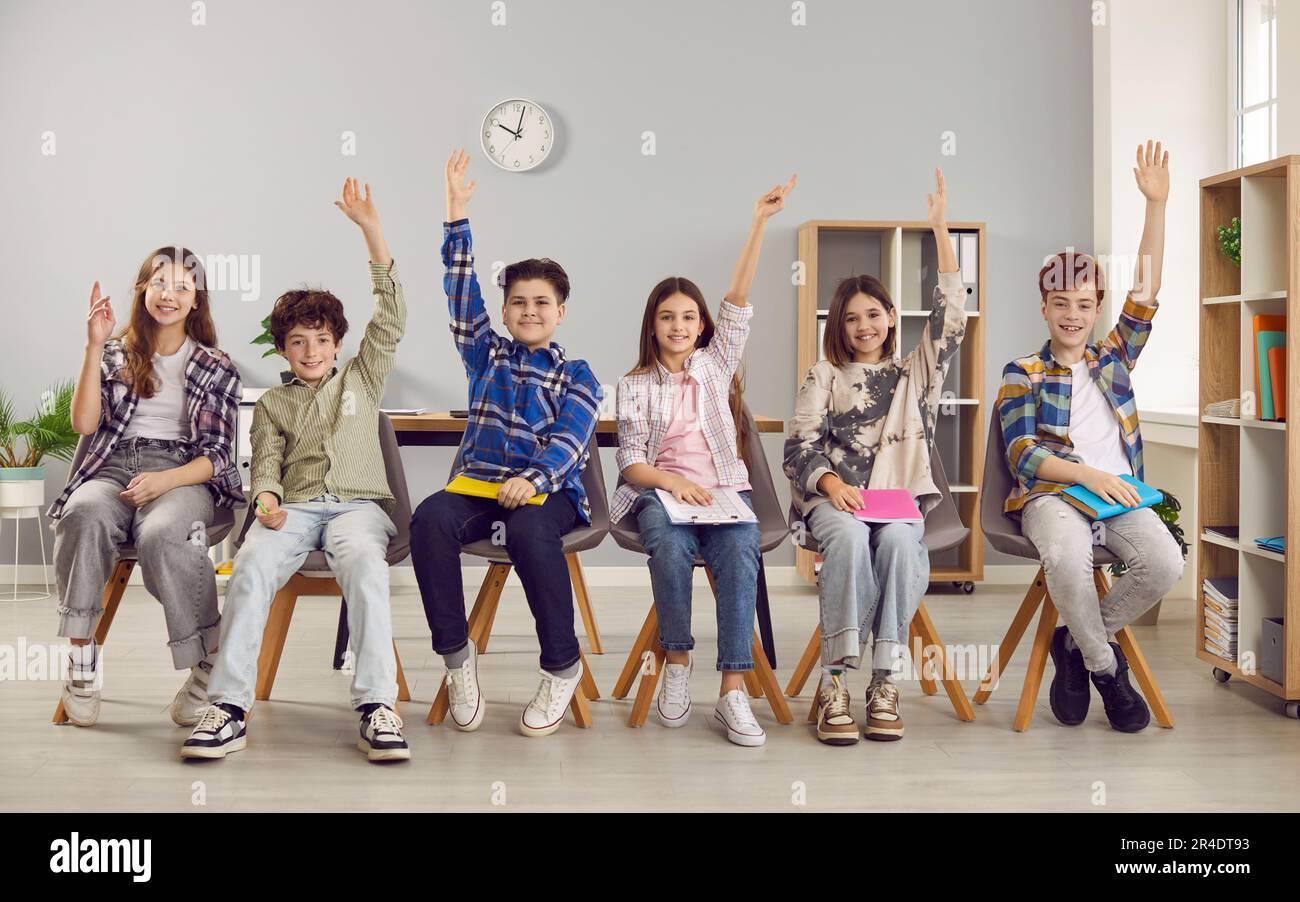 Happy school students sitting in row and raising their hands to answer ...