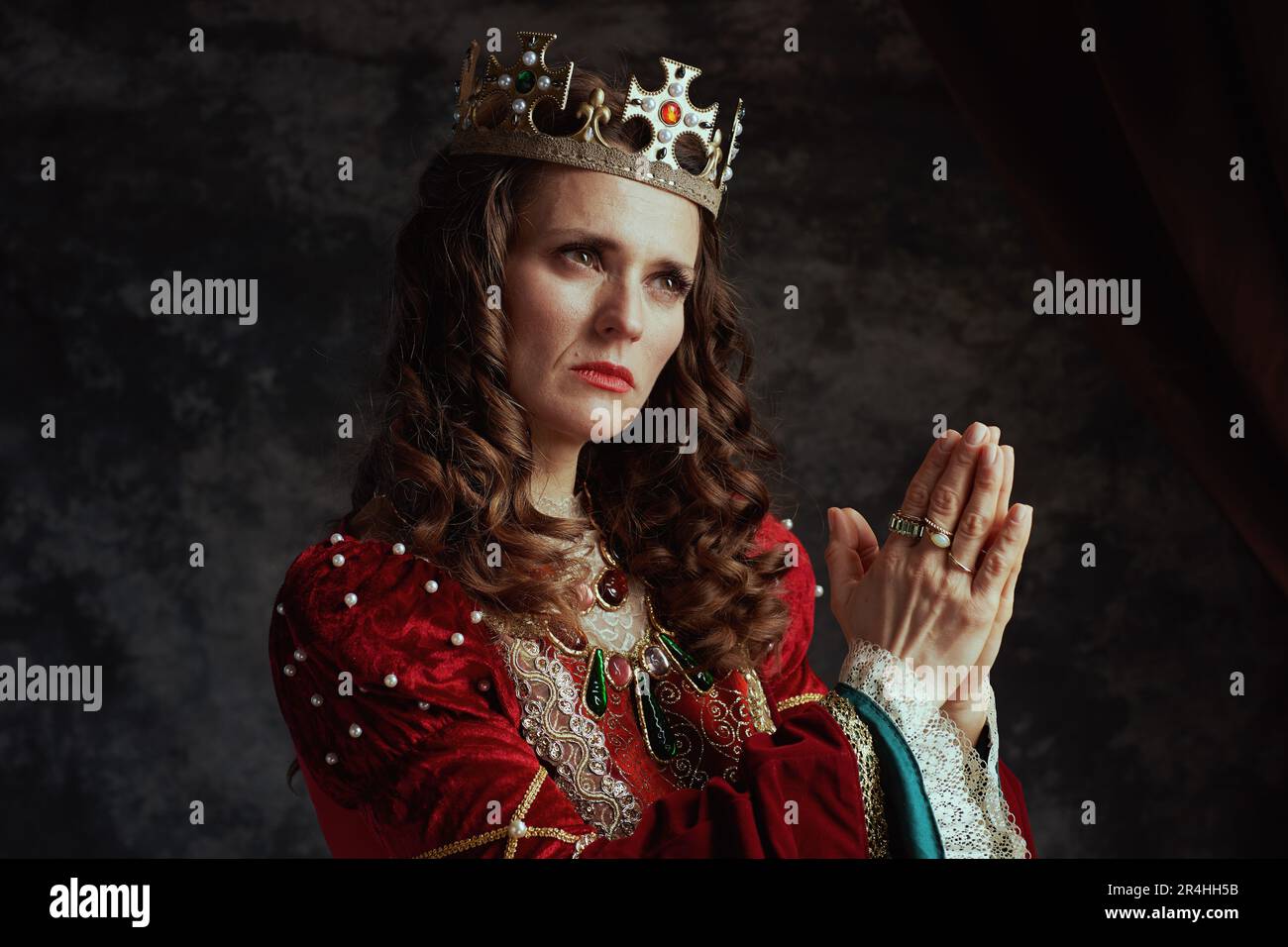 medieval queen in red dress with crown praying on dark gray background ...