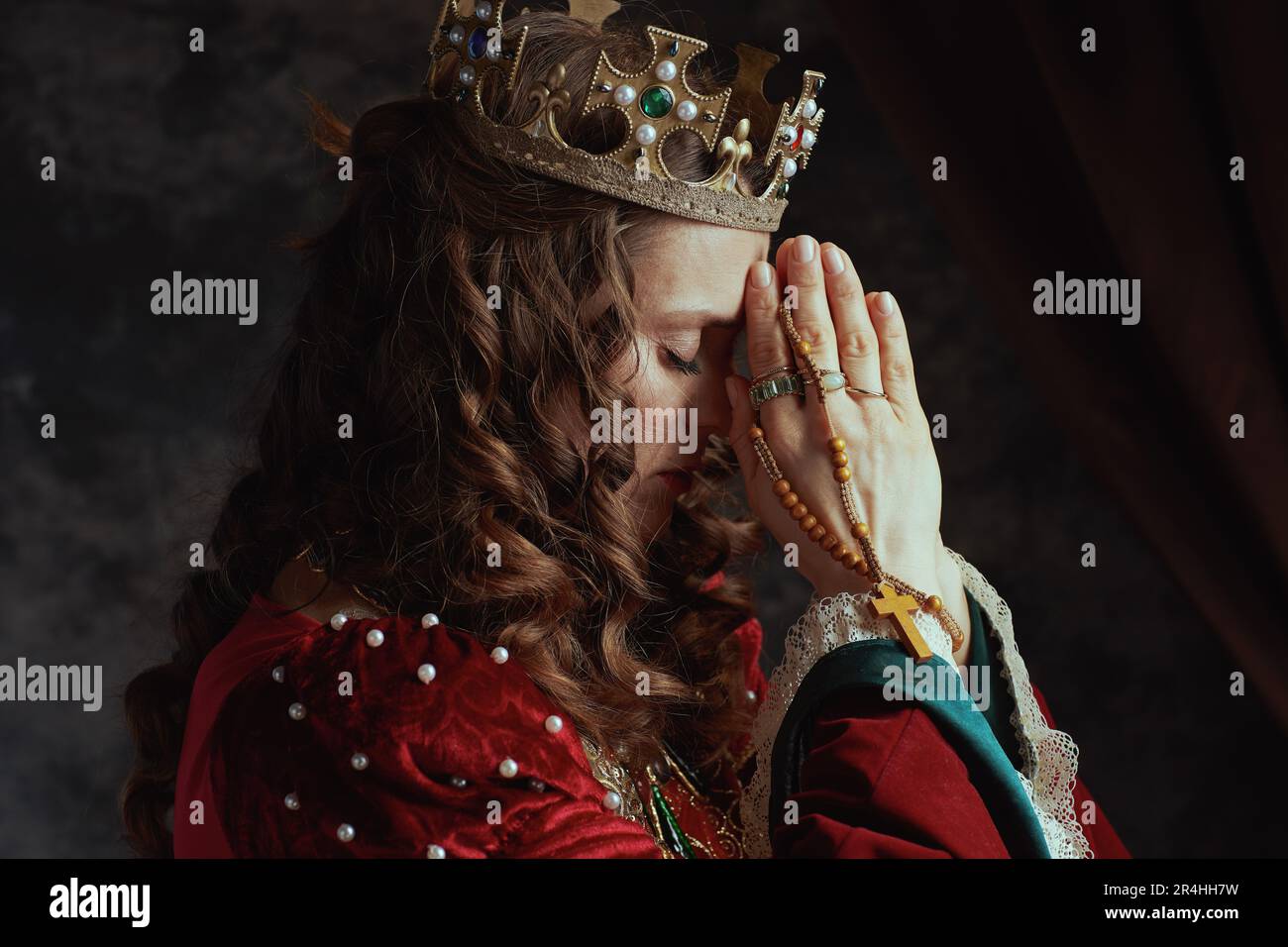 medieval queen in red dress with rosary and crown praying on dark gray ...