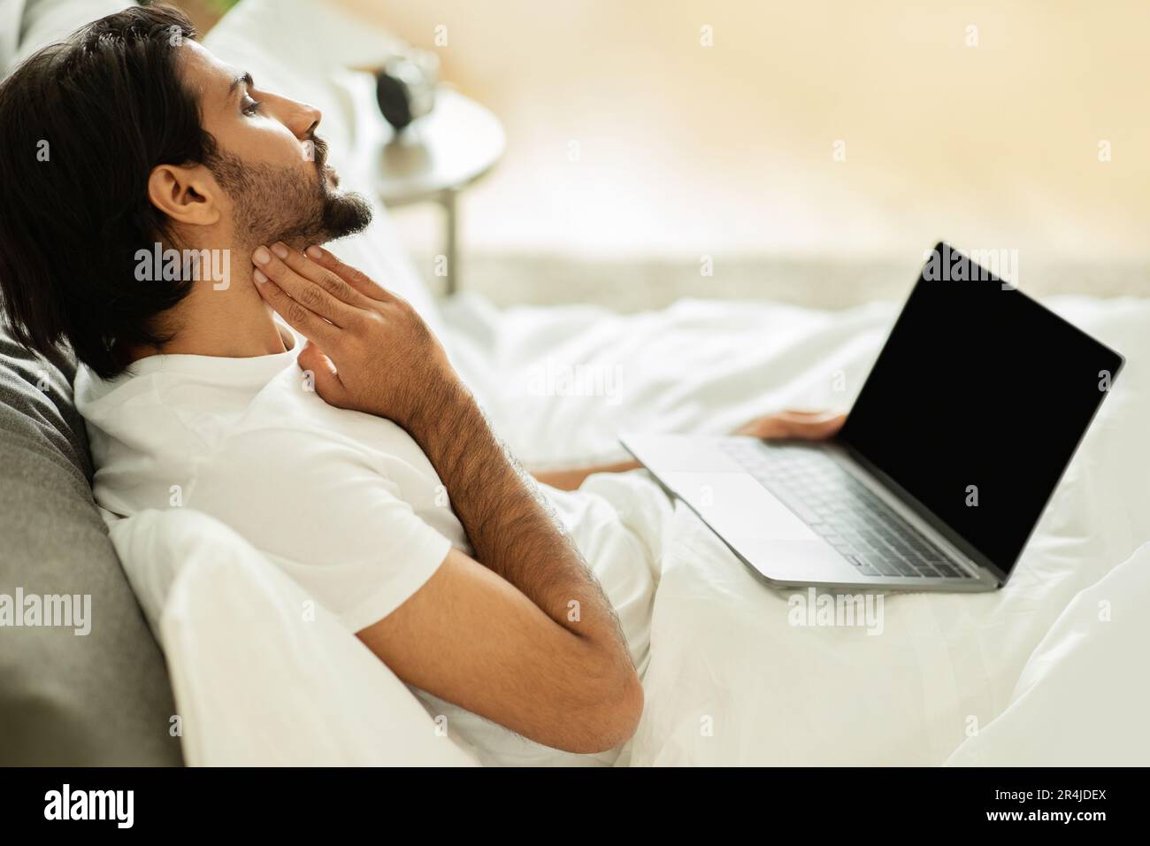 Sad middle eastern guy woke up, sits on bed, check tonsils with laptop ...
