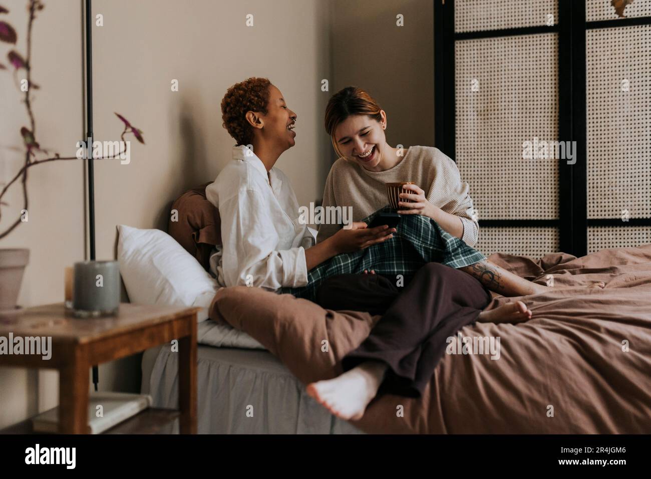 Happy non-binary couple sharing smart phone while sitting on bed at ...