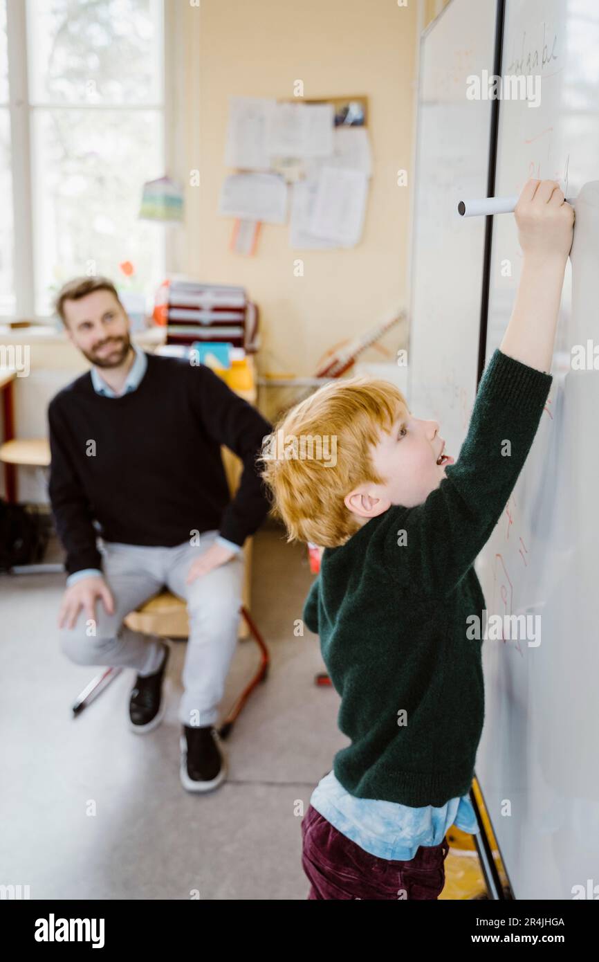 Blond boy writing on whiteboard near male teacher sitting in classroom ...