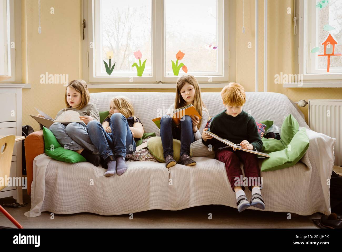 Full length of girls and boy reading books while sitting on sofa in ...