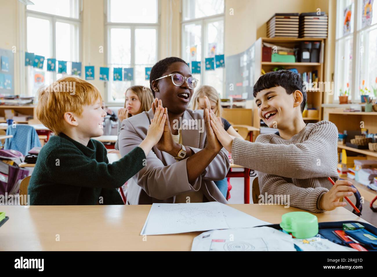 Happy teacher and boys giving high-fives while sitting at desk in ...