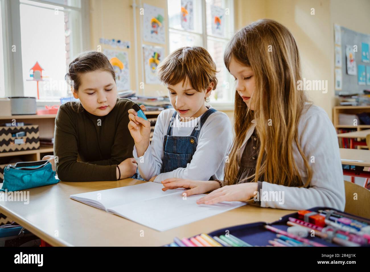 Boy and girls sharing book while studying together at desk in classroom ...