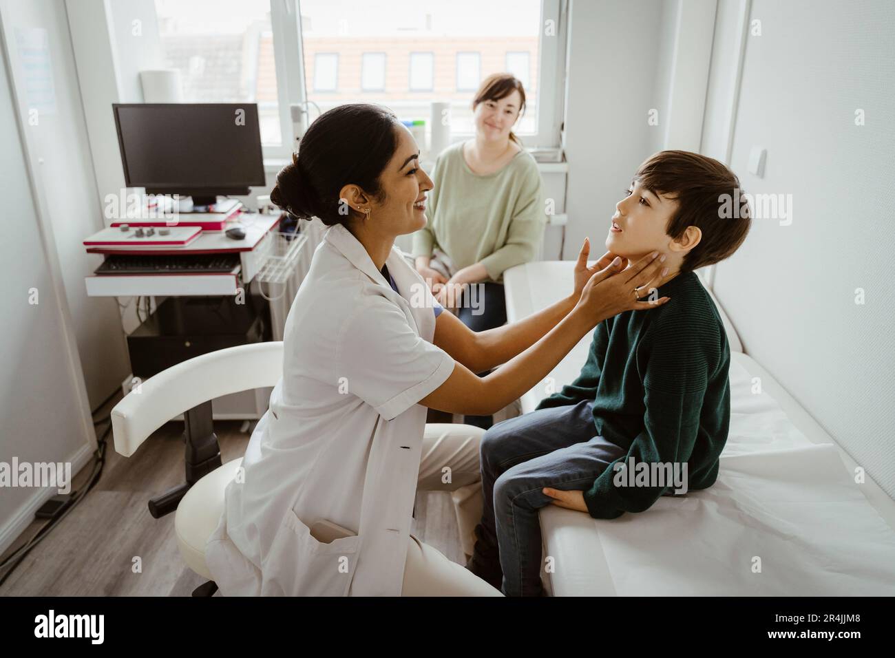 Side view of smiling female pediatrician examining boy sitting on bed ...