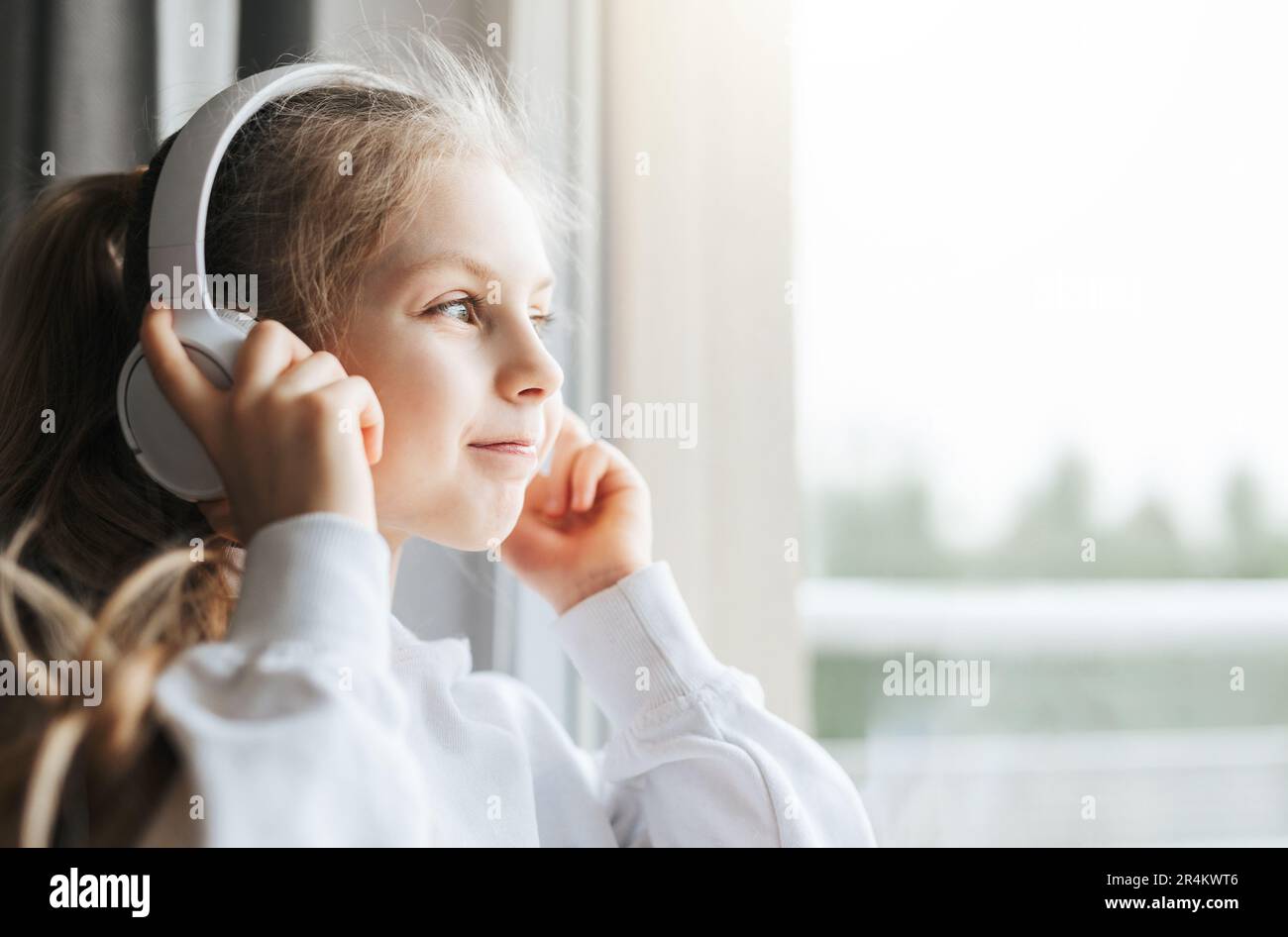 Little girl with headphones indoors at home, sitting on window sill and ...