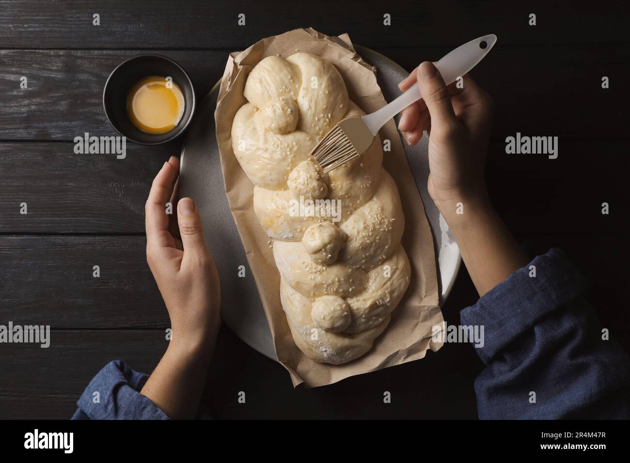 Woman spreading egg yolk onto raw braided bread at black wooden table ...