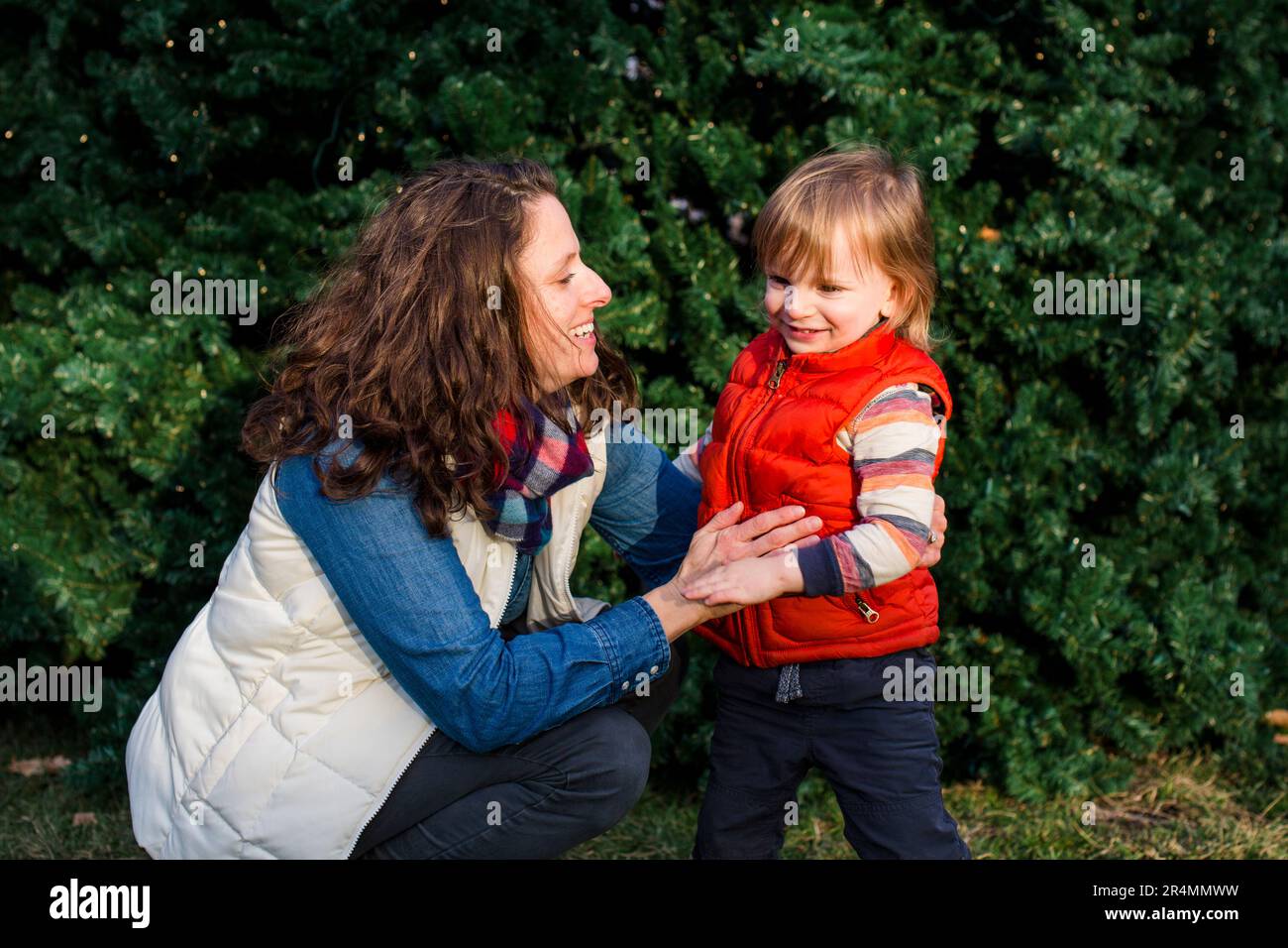 A laughing mother and small child sit together in front of lit-up tree ...