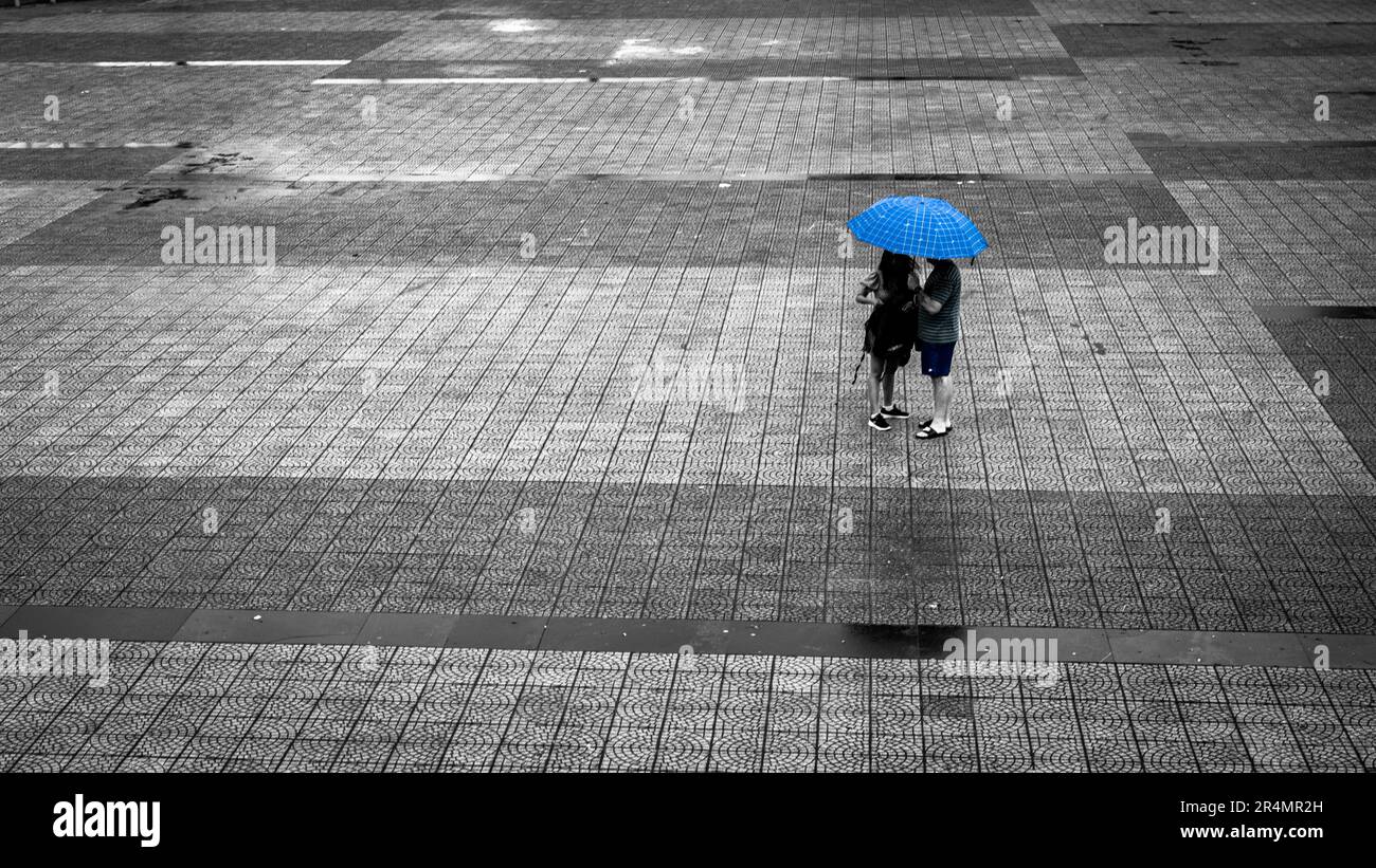 A couple shelters under a blue umbrella in the rain in the middle of an open paved area in ...