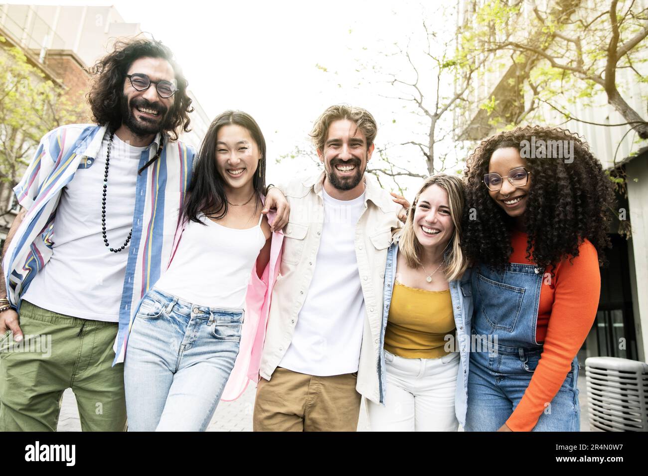 Diverse group of cheerful millennial friends standing together hugging each other in street ...