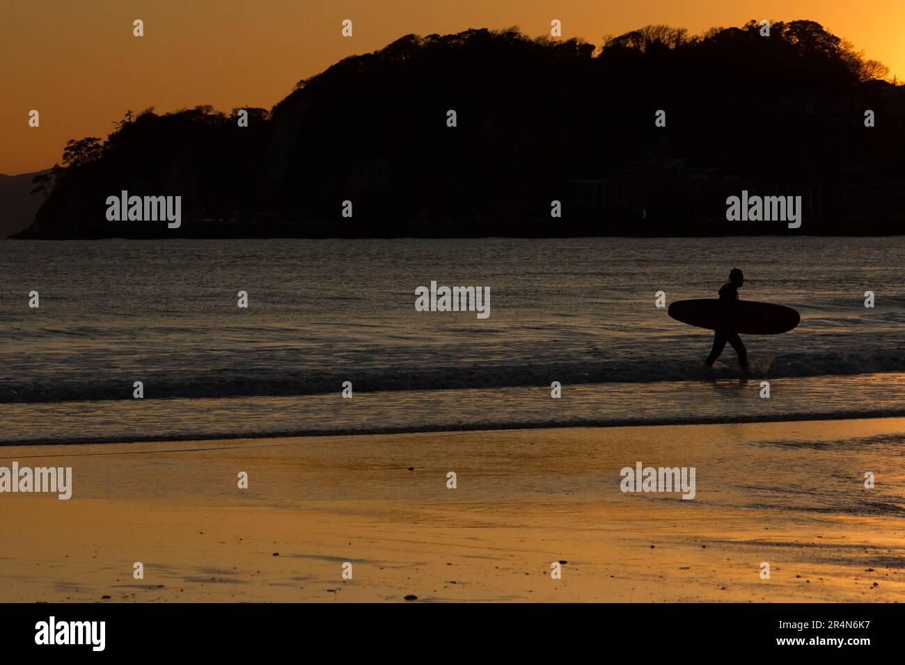 A surfer in silhouette carries a surfboard out of the ocean in front of ...