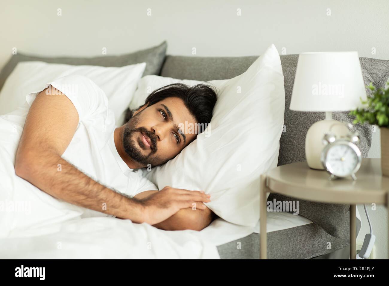 Sad serious middle eastern man woke up, lies on bed, looks at alarm ...