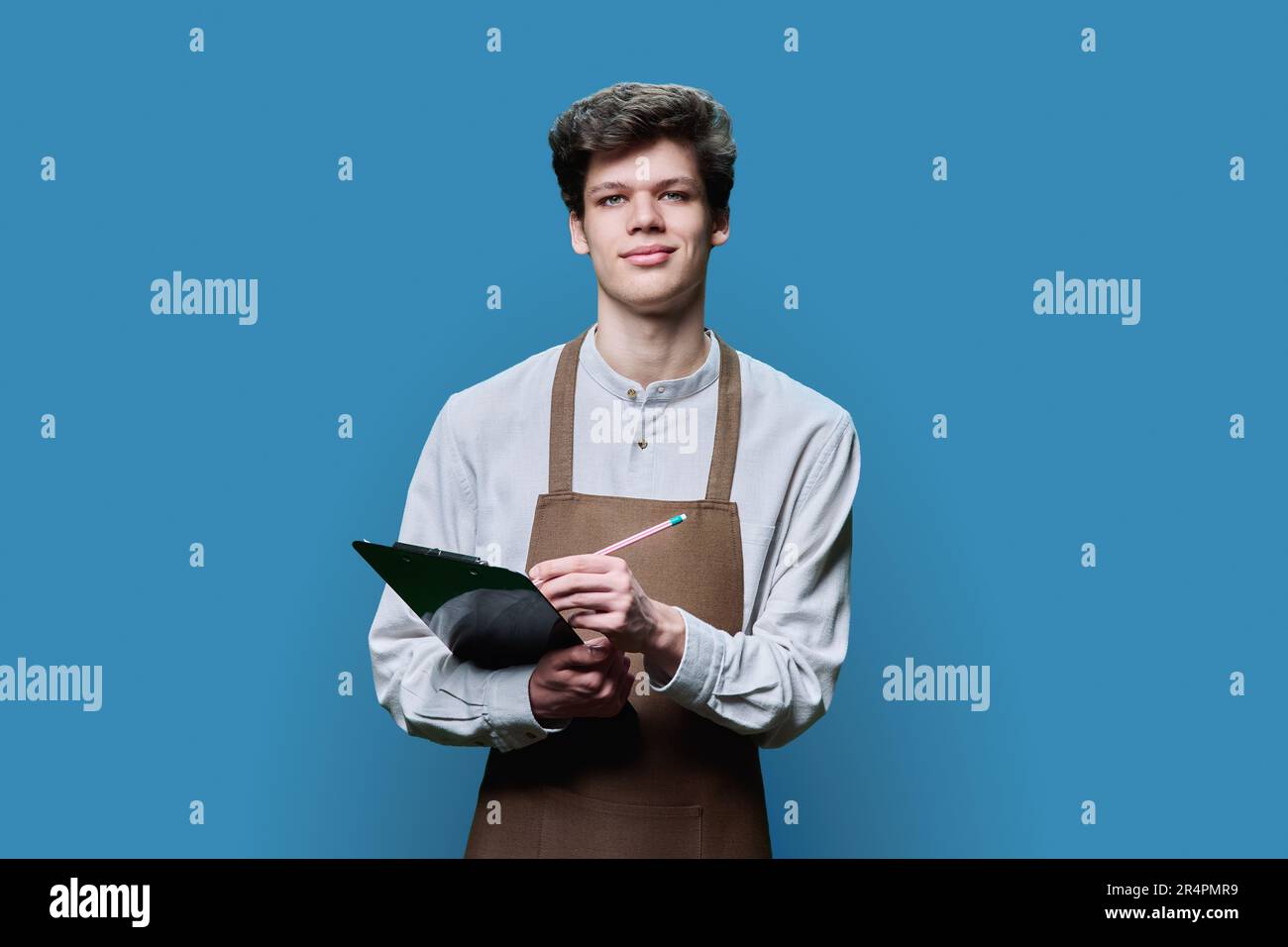 Young guy in apron making notes on clipboard, on blue studio background ...