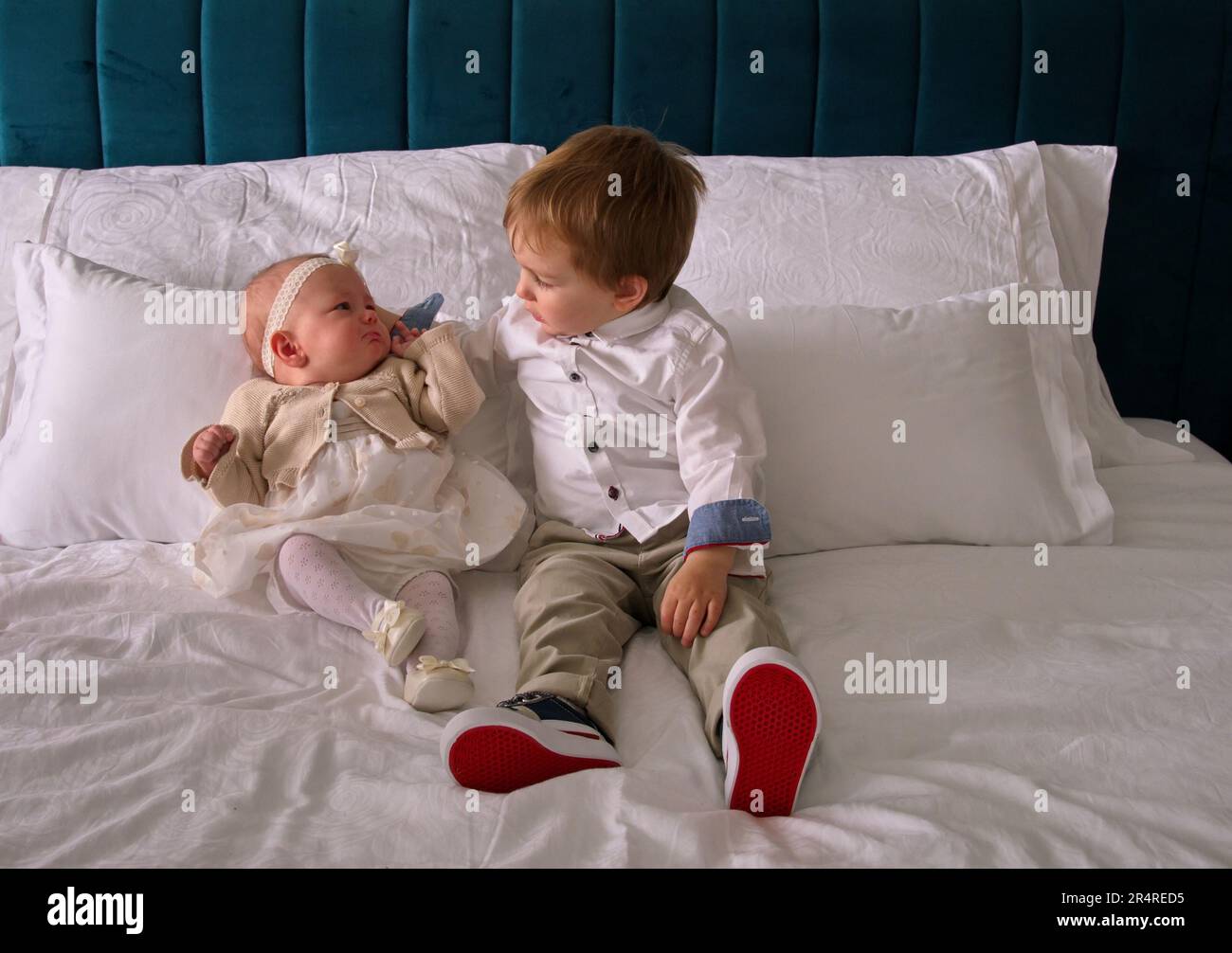 Toddler and his newborn baby sister sitting on a bed dressed for ...