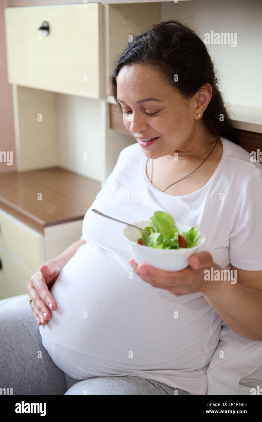 Smiling pregnant woman caressing her belly, eating healthy salad ...