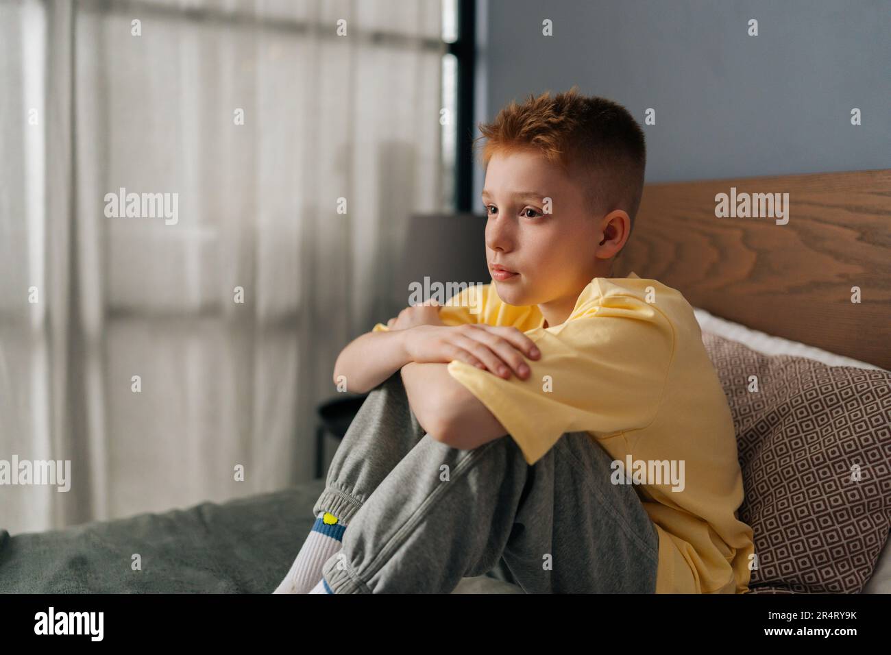 Scared sad little boy sitting on bed alone with hugging knee and ...