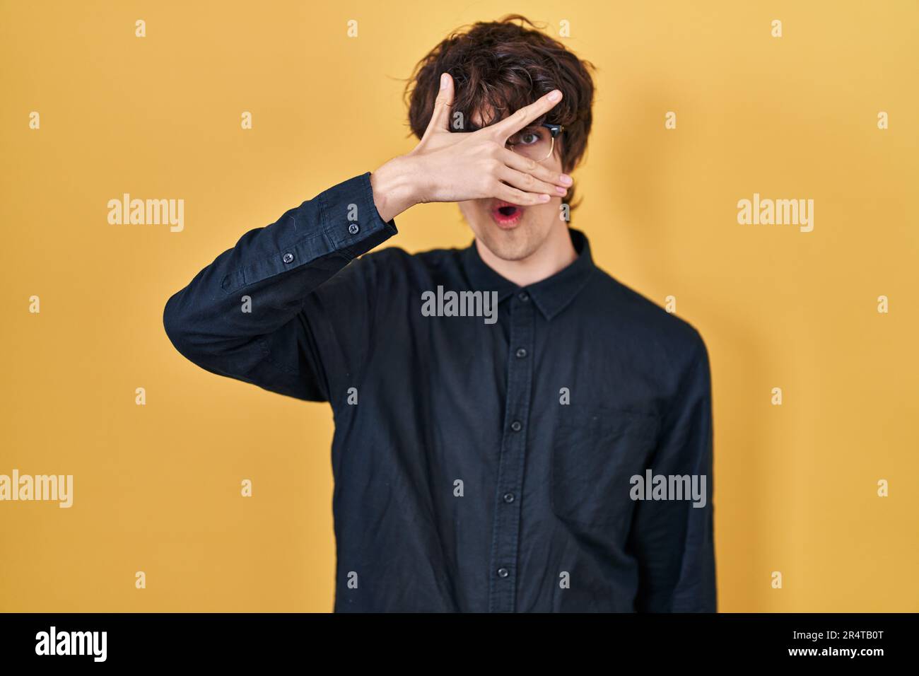Young man wearing glasses over yellow background peeking in shock ...