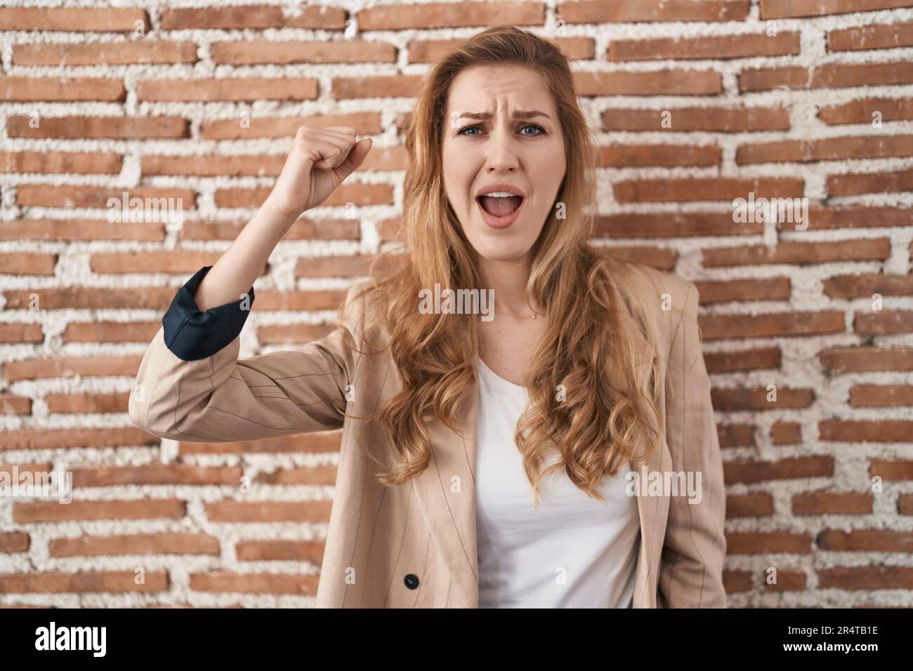 Beautiful blonde woman standing over bricks wall angry and mad raising ...