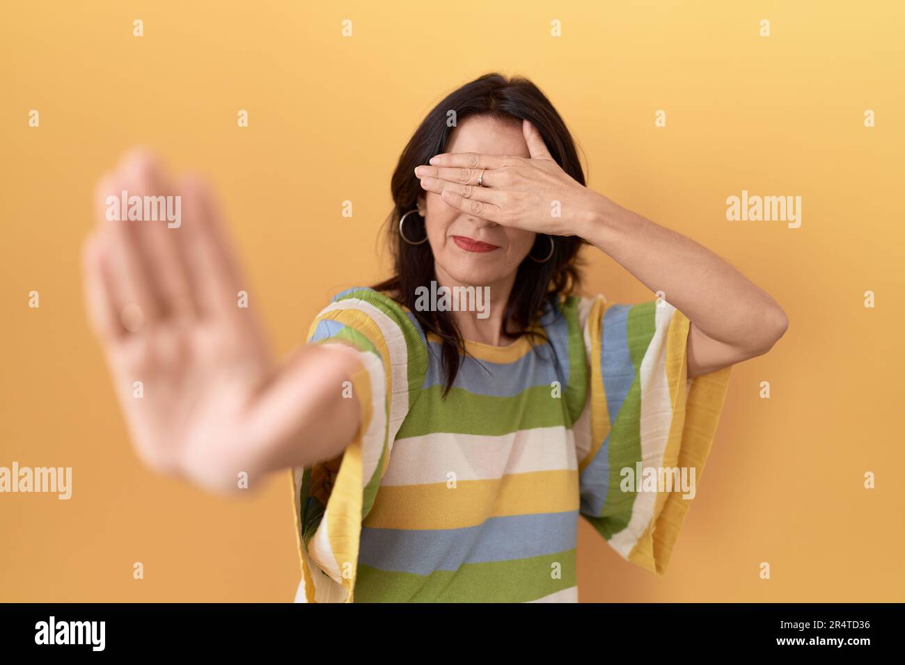 Middle age hispanic woman standing over yellow background covering eyes ...