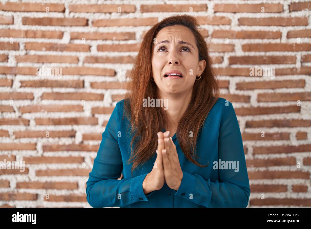 Brunette woman standing over bricks wall begging and praying with hands ...