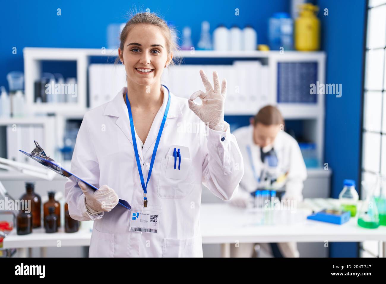 Blonde woman working at scientist laboratory doing ok sign with fingers ...