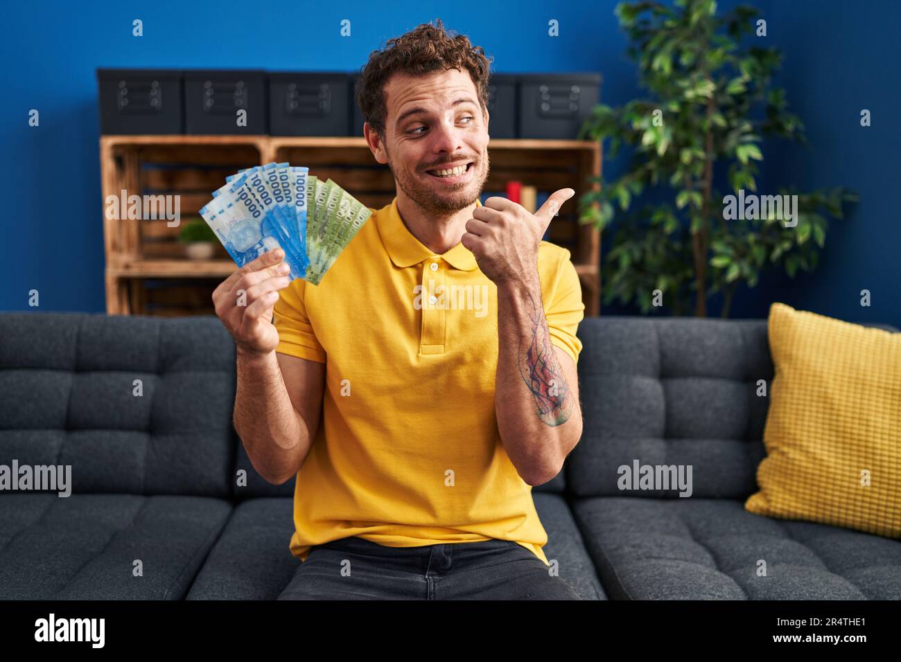 Young hispanic man holding chilean pesos pointing thumb up to the side ...