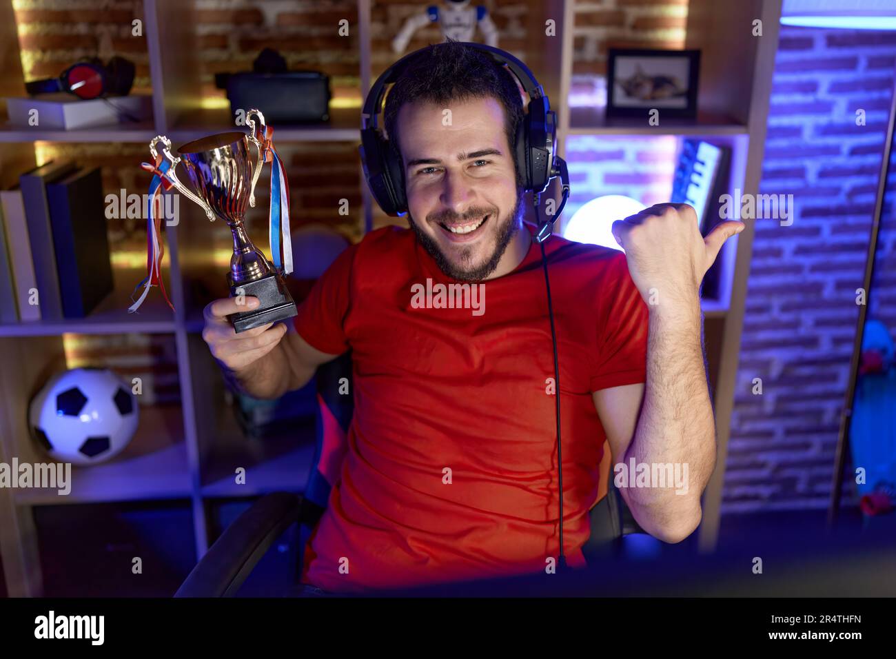 Young hispanic man playing video games holding trophy pointing thumb up ...