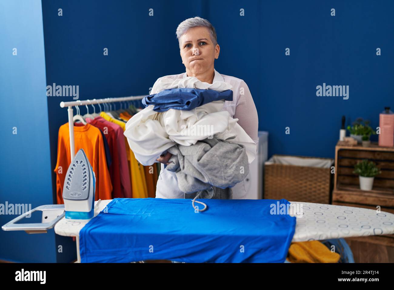Middle age woman ironing clothes at home puffing cheeks with funny face ...